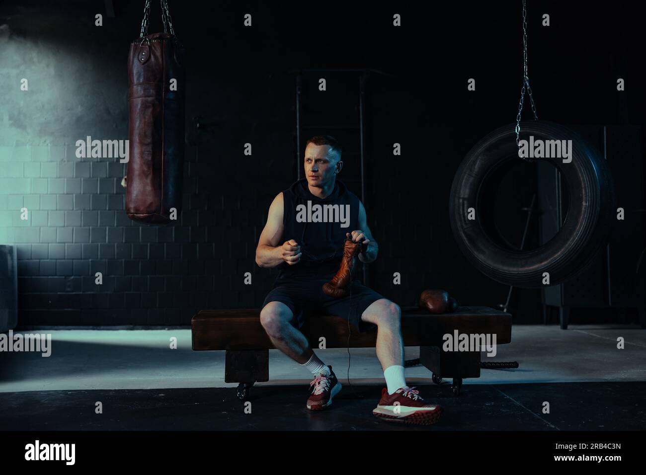 Male boxer sitting on bench in dark gym and putting on boxing gloves ...