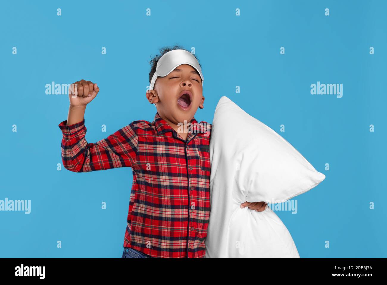 Boy with pillow and sleep mask yawning and stretching on light blue ...