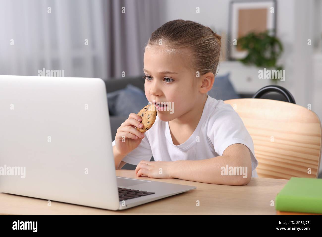 Little girl using laptop and eating biscuit at table indoors. Internet ...