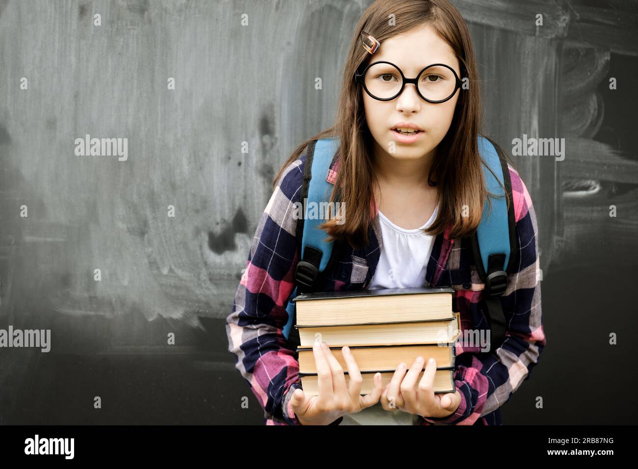 Teen girl little student holding pile of heavy books on chalkboard ...