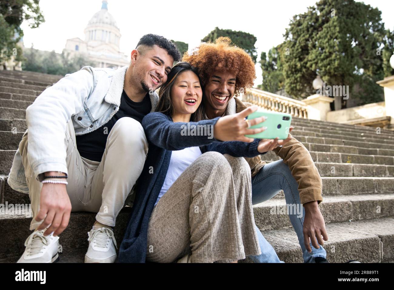 Three young diverse people taking a selfie with a smartphone sitting on a stairs. Multiracial ...