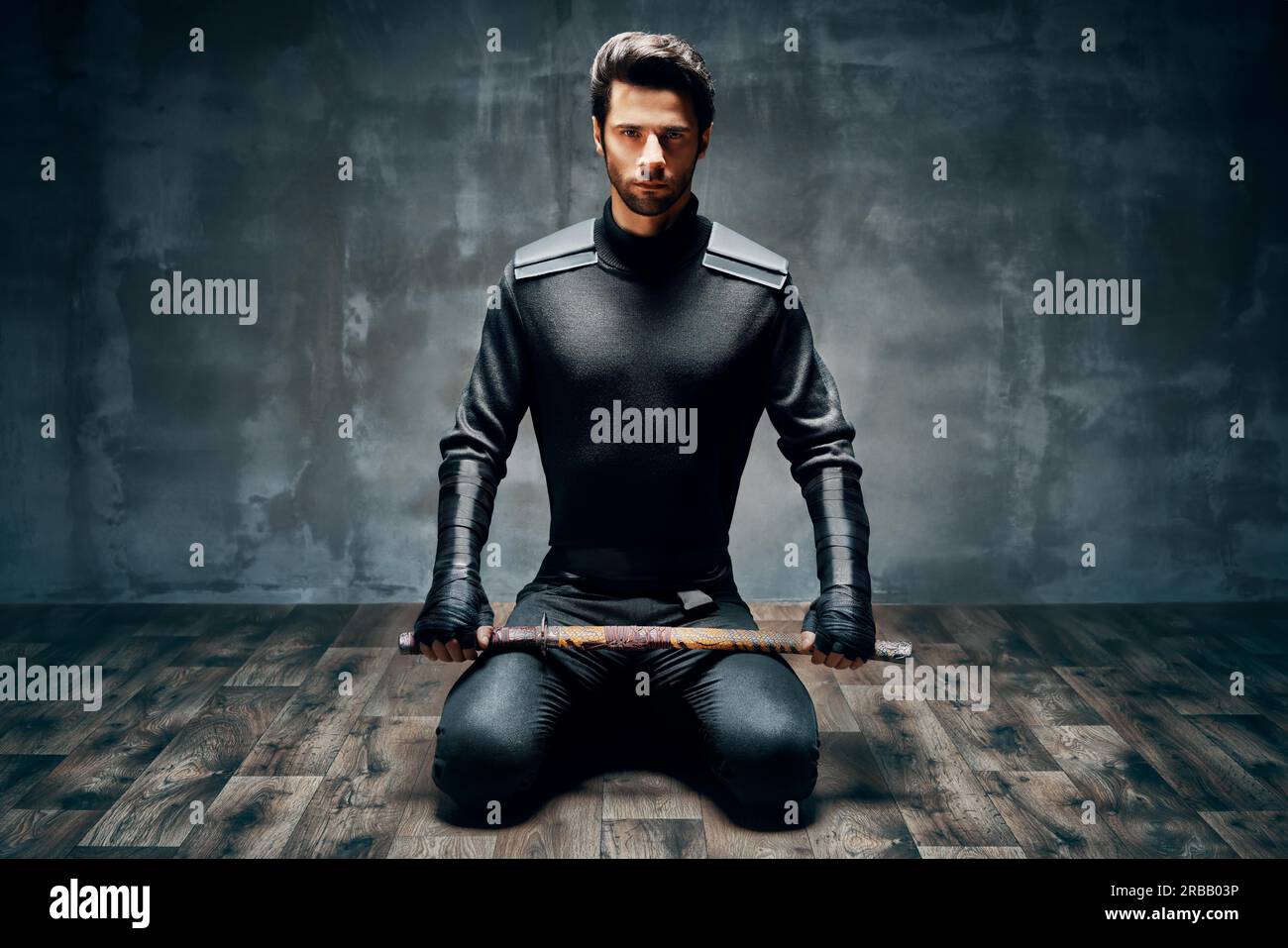 Warrior man sitting on floor posing with a sword over black background ...