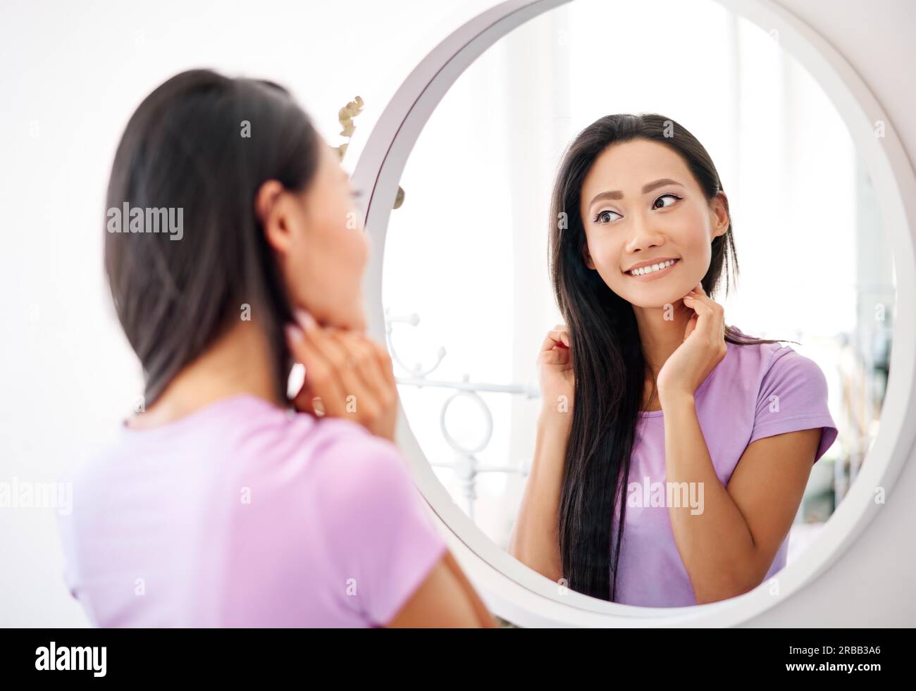 Smiling beautiful asian woman looking at herself in the mirror at home ...