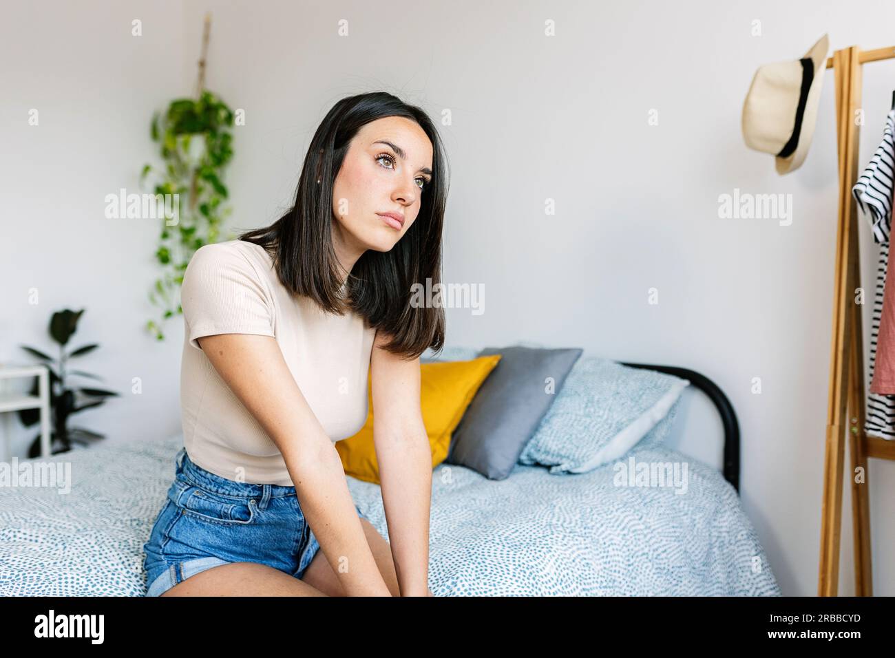 Sad young adult woman sitting on bed looking through the window at home ...
