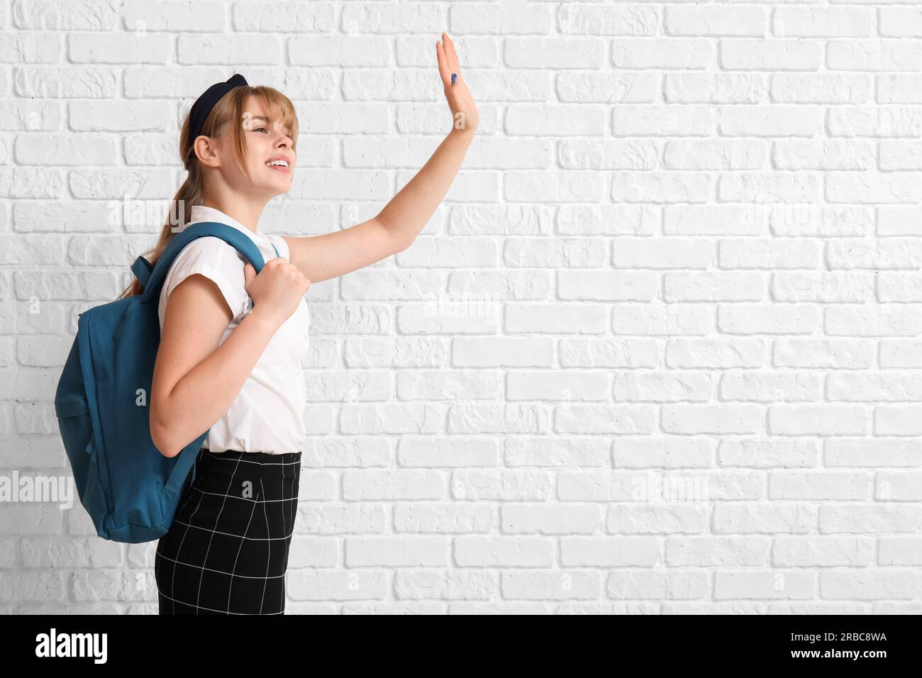 Female student with backpack waving hand on white brick background ...