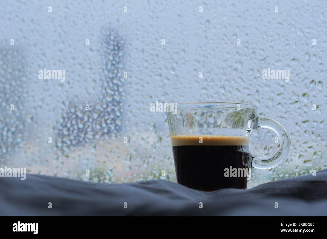 A glass of Espresso coffee shot puts on bed with rain drop on window in ...