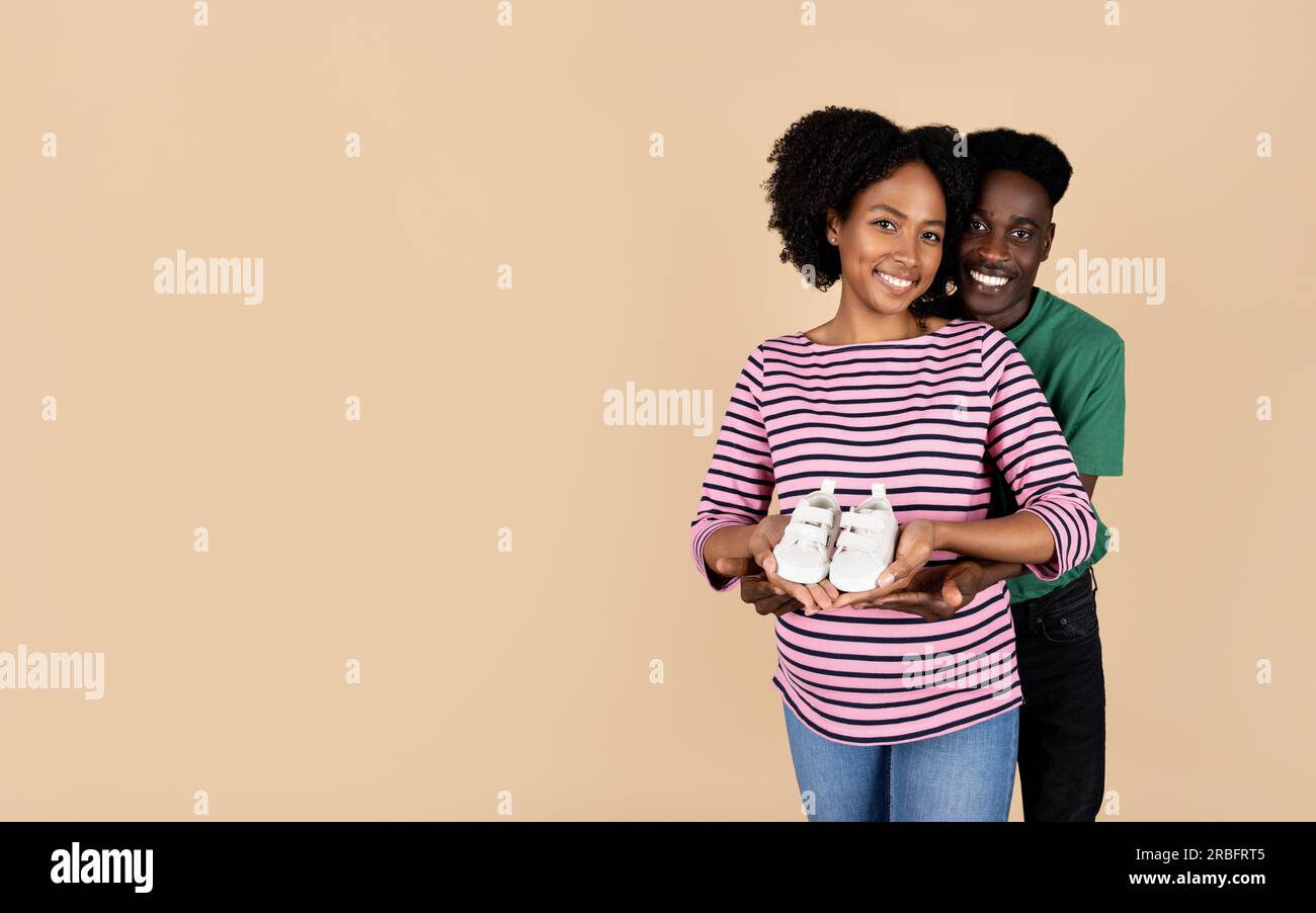 Glad millennial african american husband hug wife with big belly, show small sneakers Stock ...