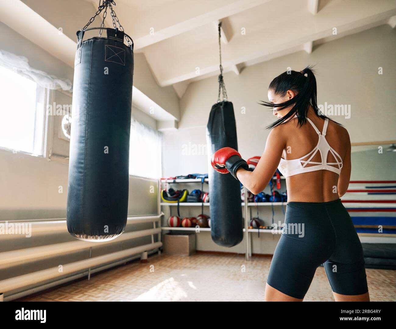 Back view of female boxer hitting a huge punching bag at fitness gym. Woman practicing her ...