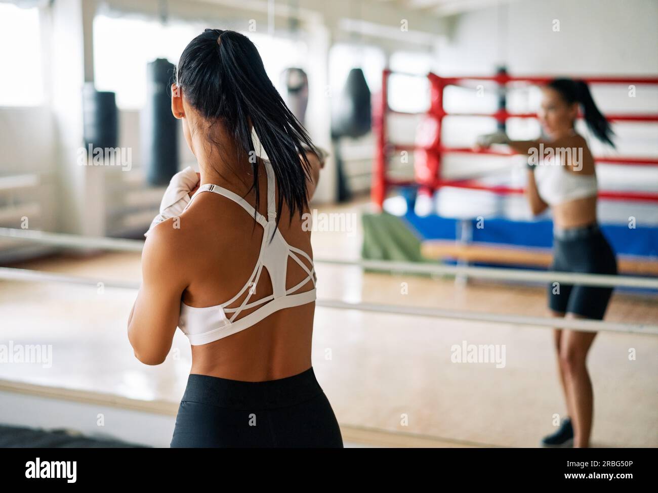 Back view of female boxer doing shadow boxing in gym. Woman practicing her punches making direct ...