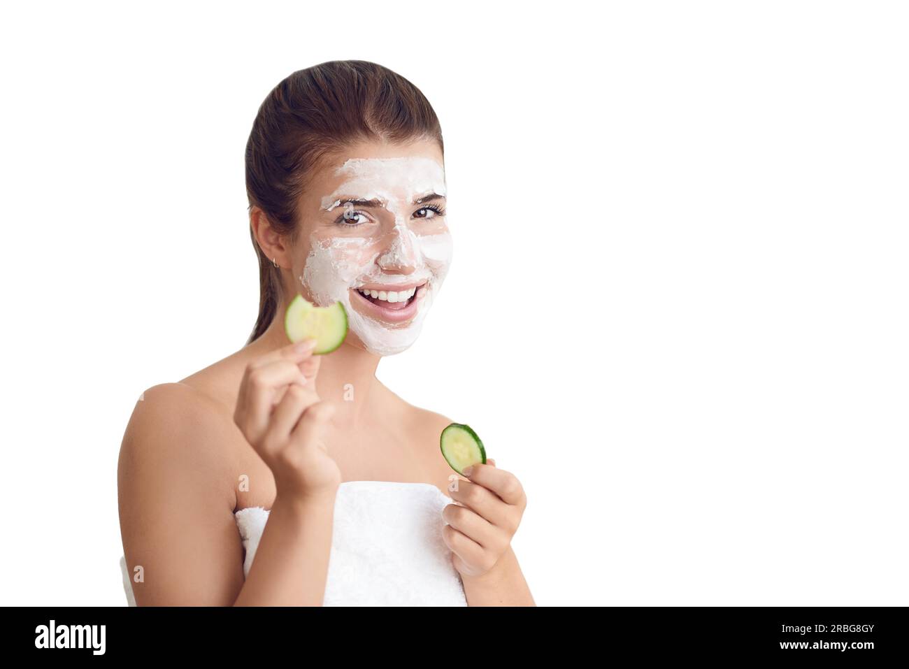 Smiling attractive young woman in a towel wearing a face mask treatment holding up slices of raw ...