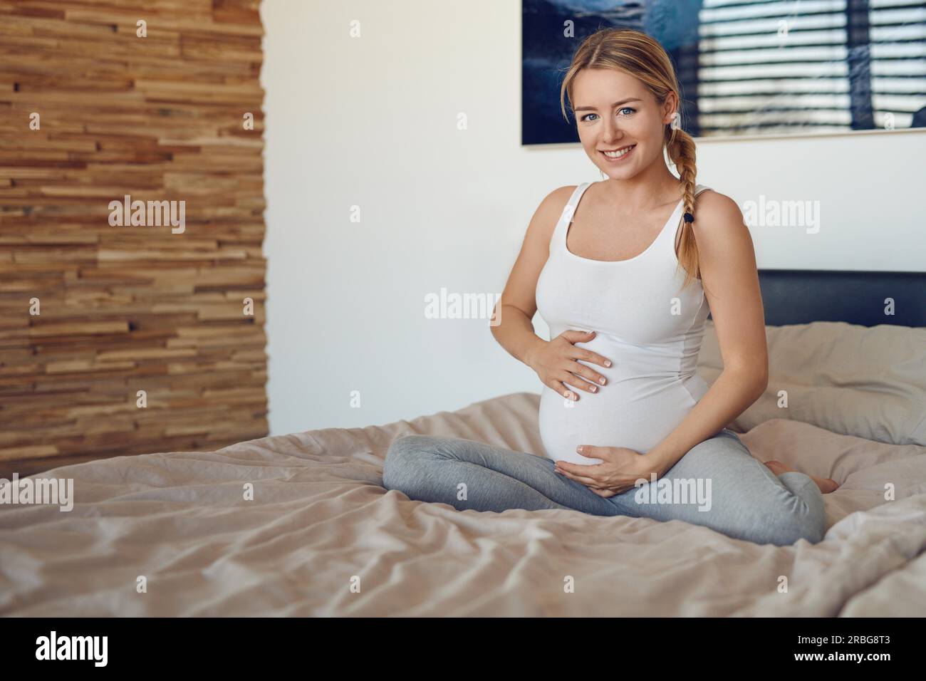 Happy pregnant woman sitting on a bed cradling her baby bump with her ...