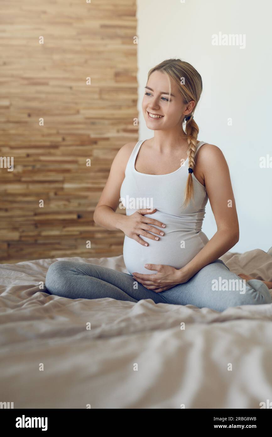 Happy young pregnant woman bonding with her unborn child sitting on a bed cradling her baby bump ...