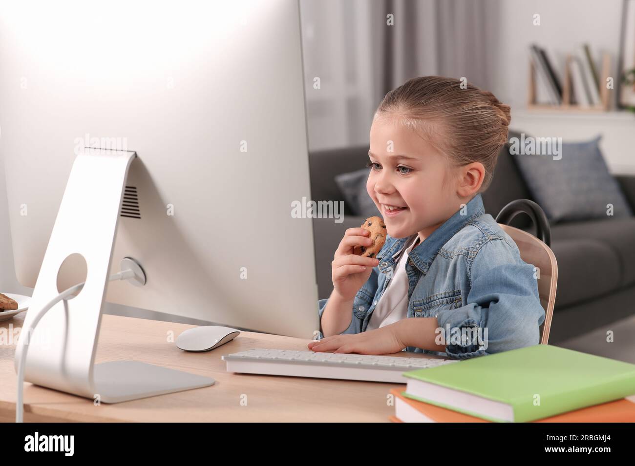 Little girl eating biscuit while using computer at table in room ...