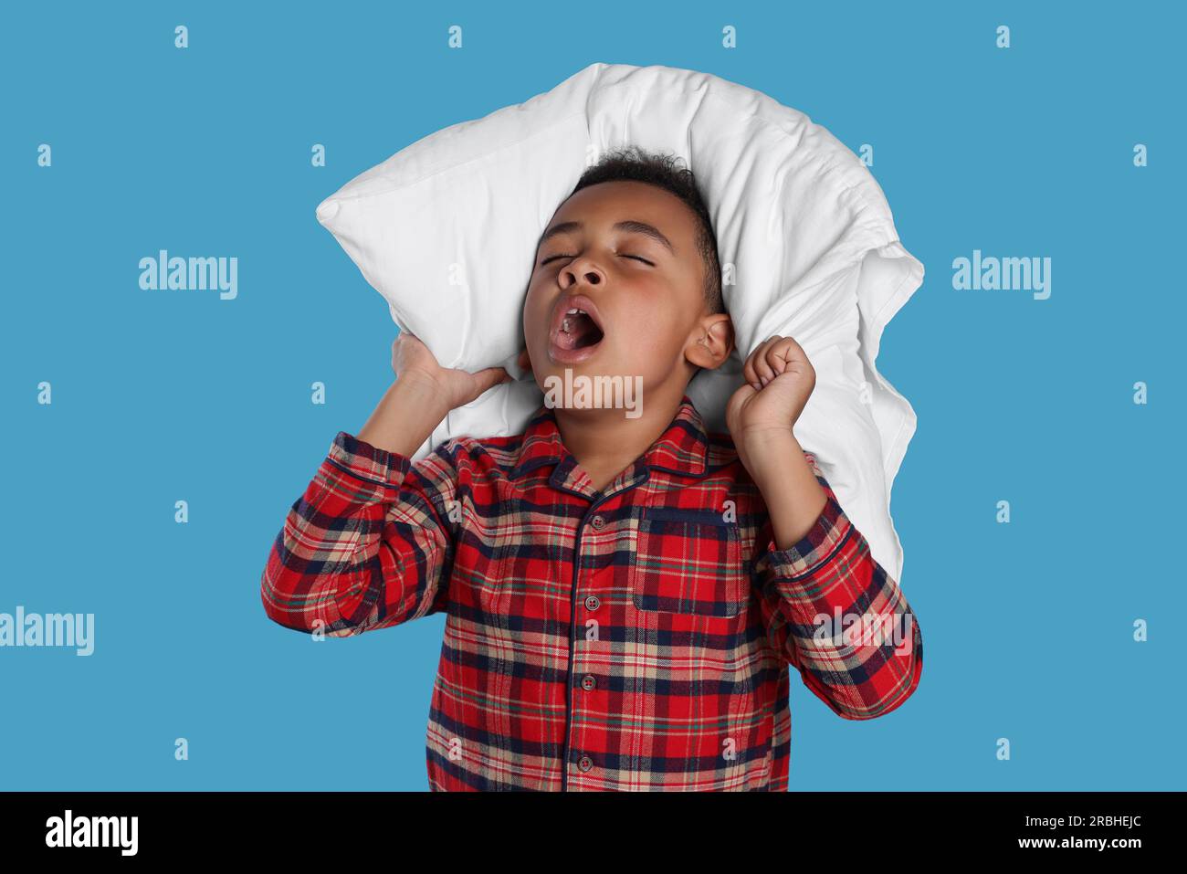 Boy covering his ears with pillow and yawning on light blue background ...