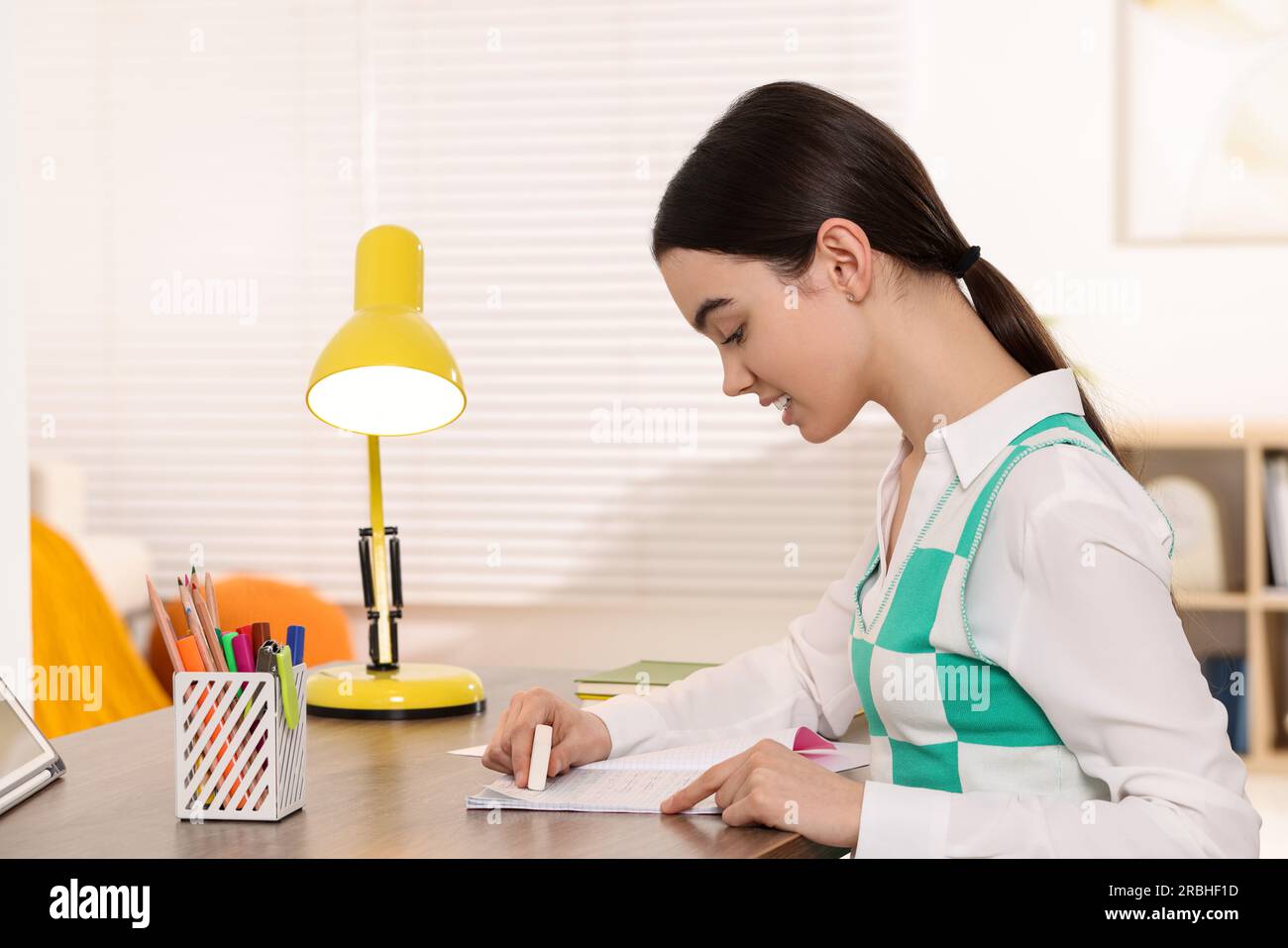 Teenage girl erasing mistake in her notebook at wooden desk indoors ...