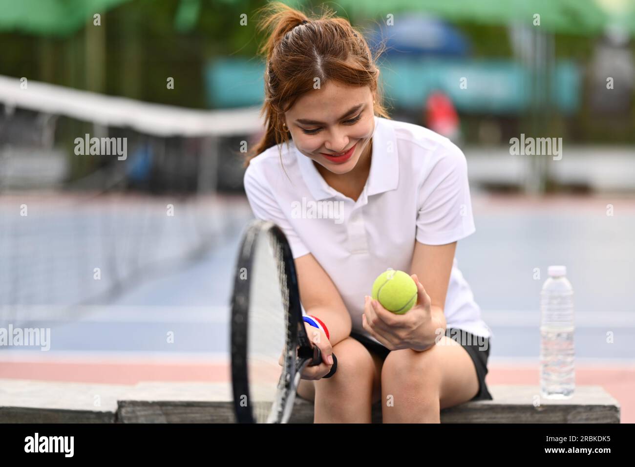 Pretty young sportswoman holding ball racket sitting on the bench at ...