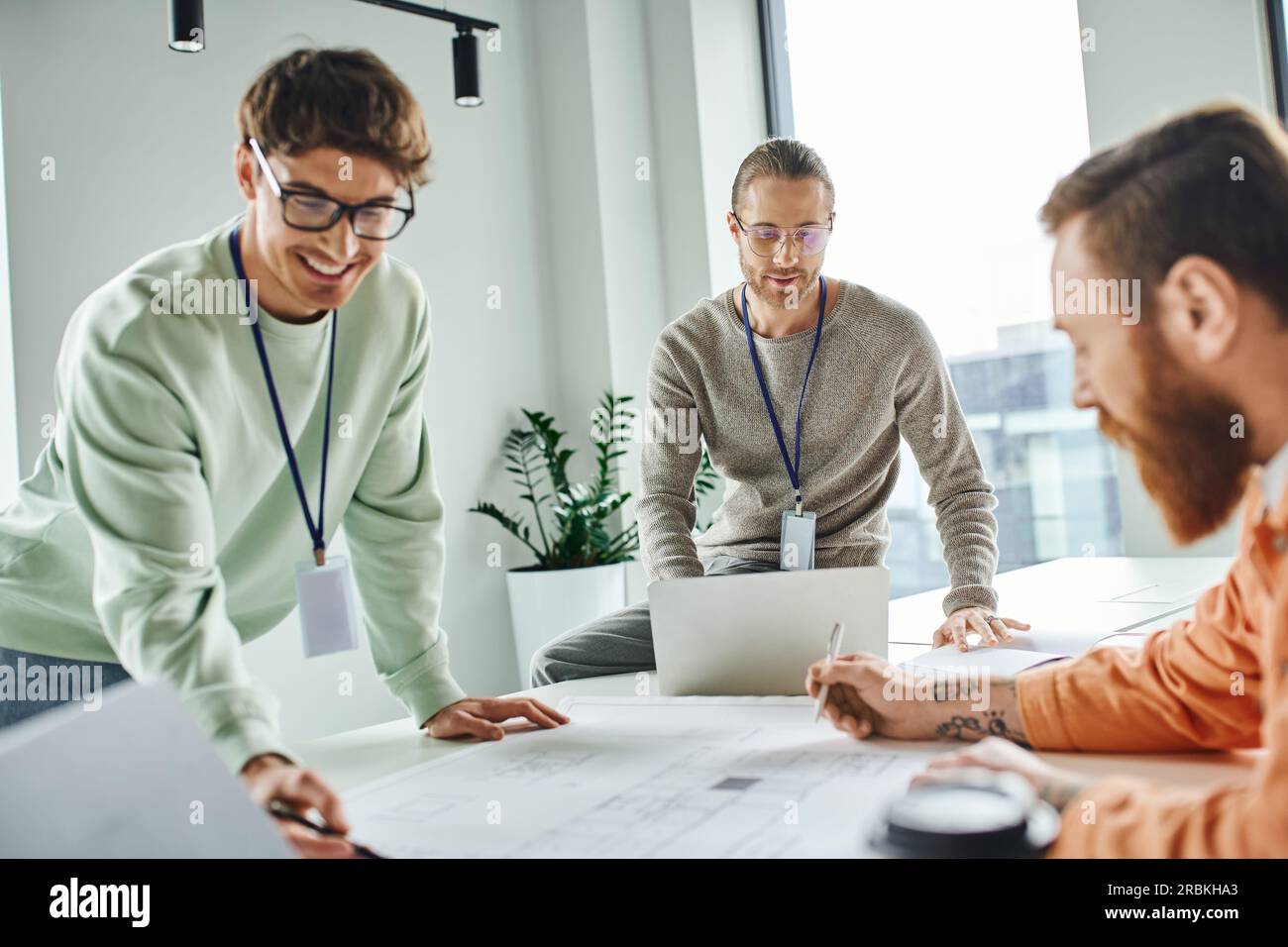 smiling architect pointing at blueprint near colleagues and laptop ...