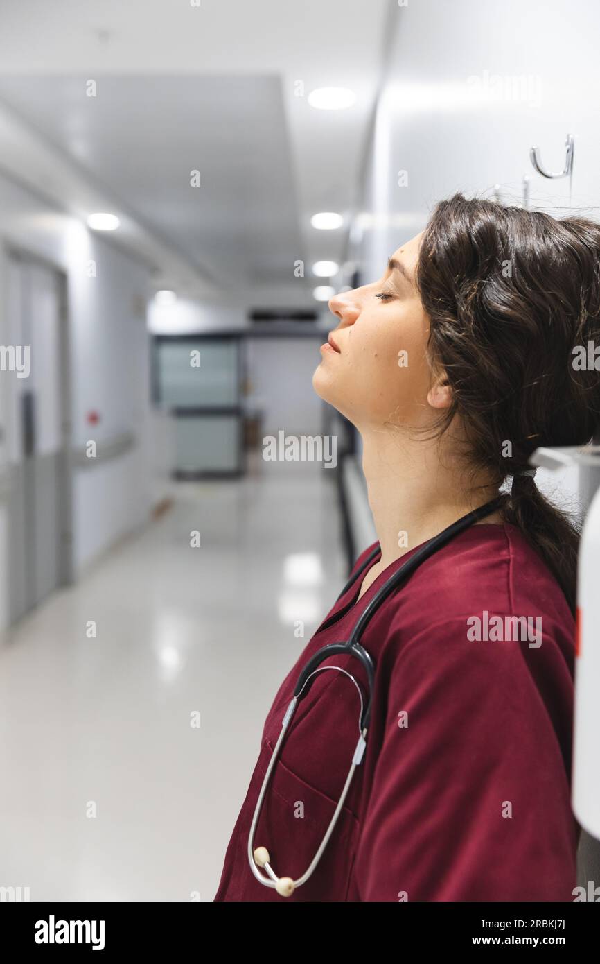 Tired caucasian female doctor wearing scrubs, leaning on wall in ...