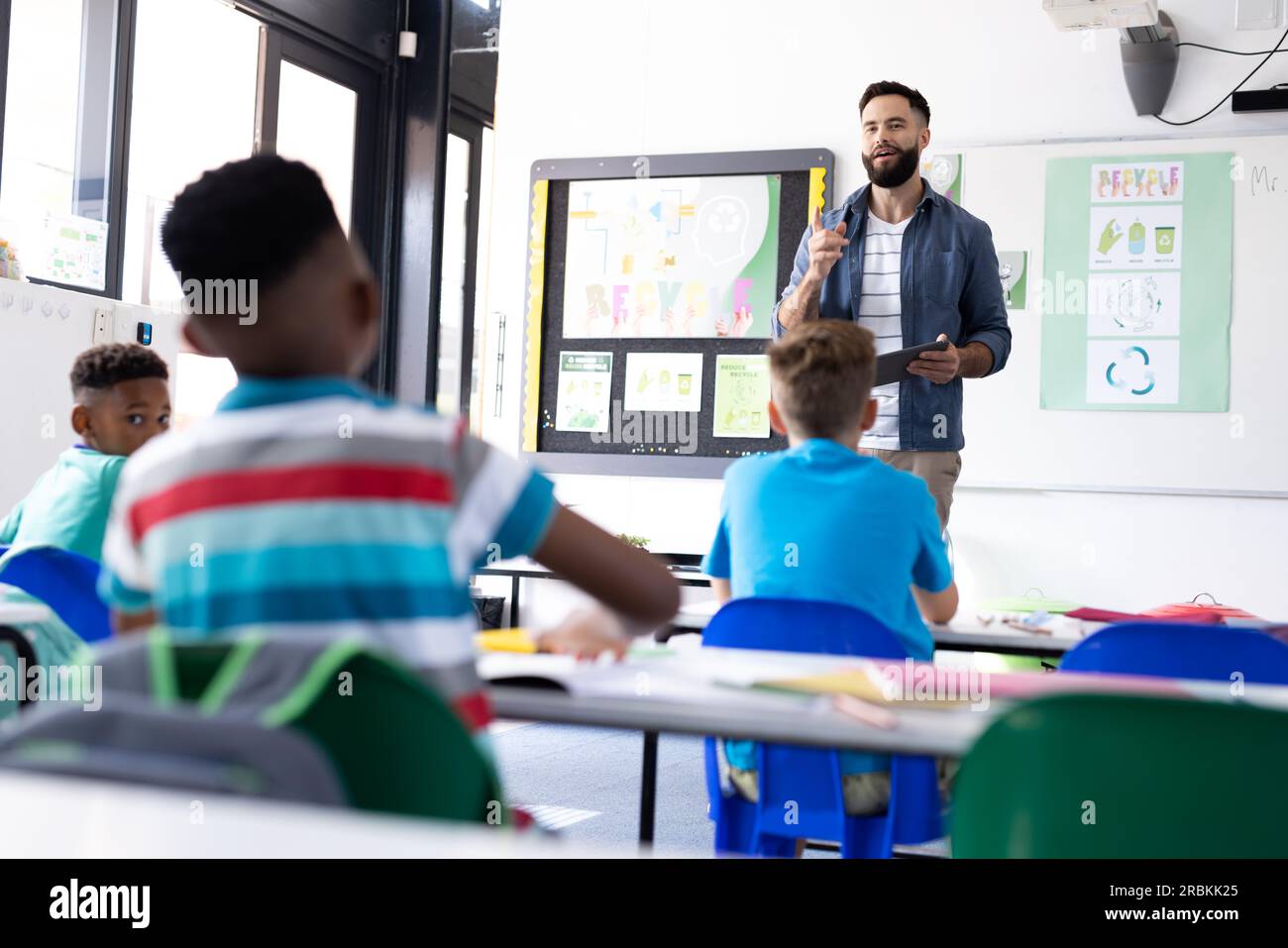 Diverse male teacher talking to pupils at desks in elementary school ...