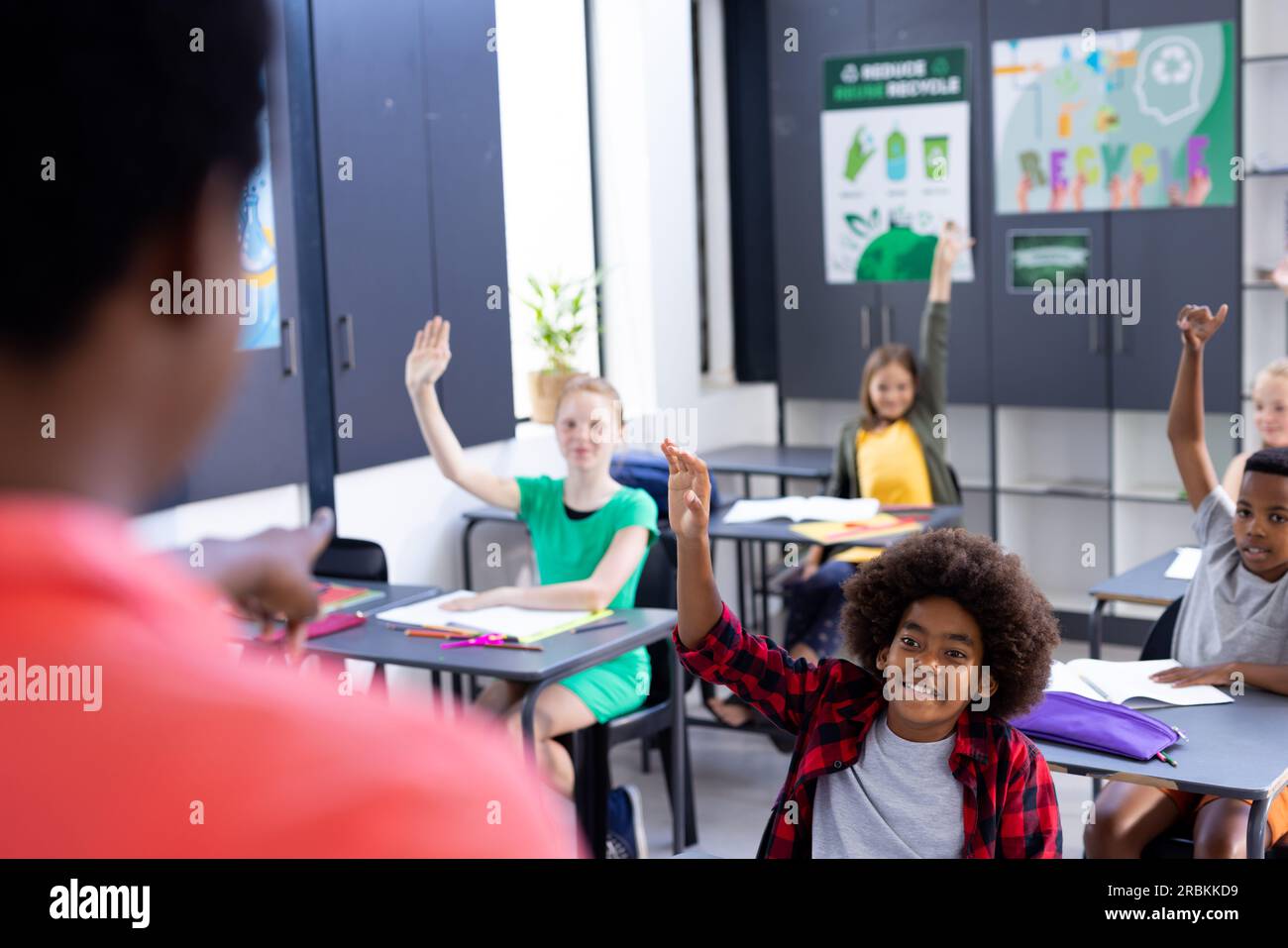 Diverse female teacher and pupils sitting at desks raising hands in ...