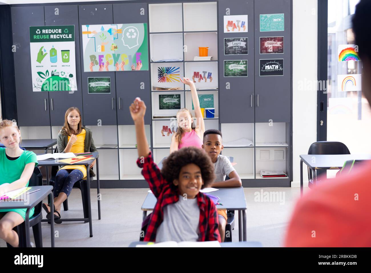 Diverse female teacher and pupils sitting at desks raising hands in ...