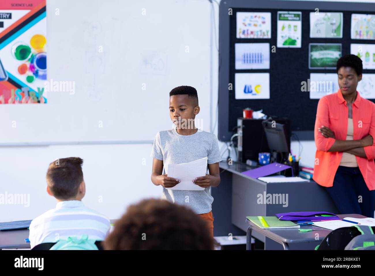 Diverse female teacher and pupils sitting at desks listening to ...