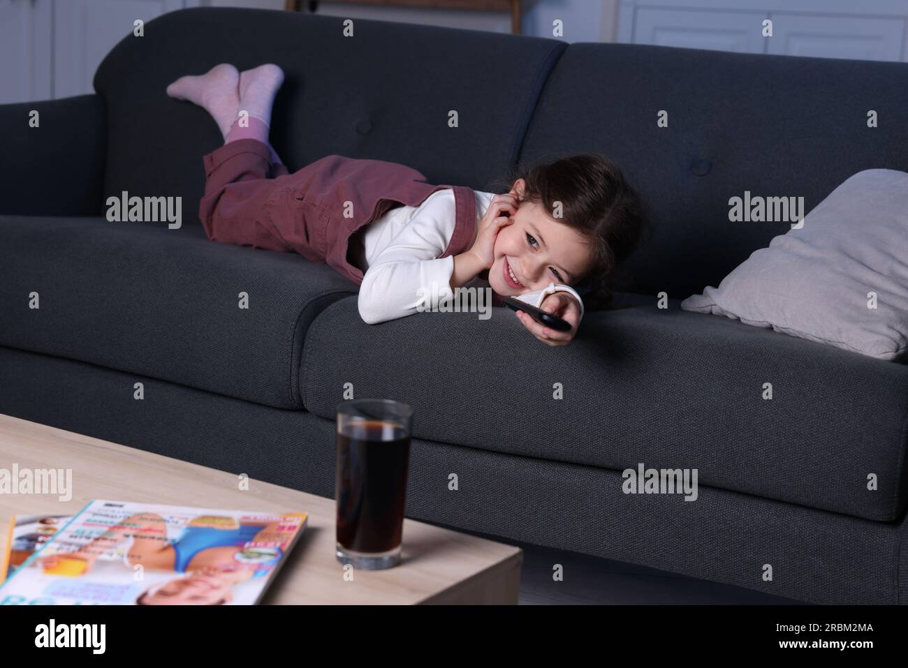Cute little girl changing TV channels with remote control on sofa at ...