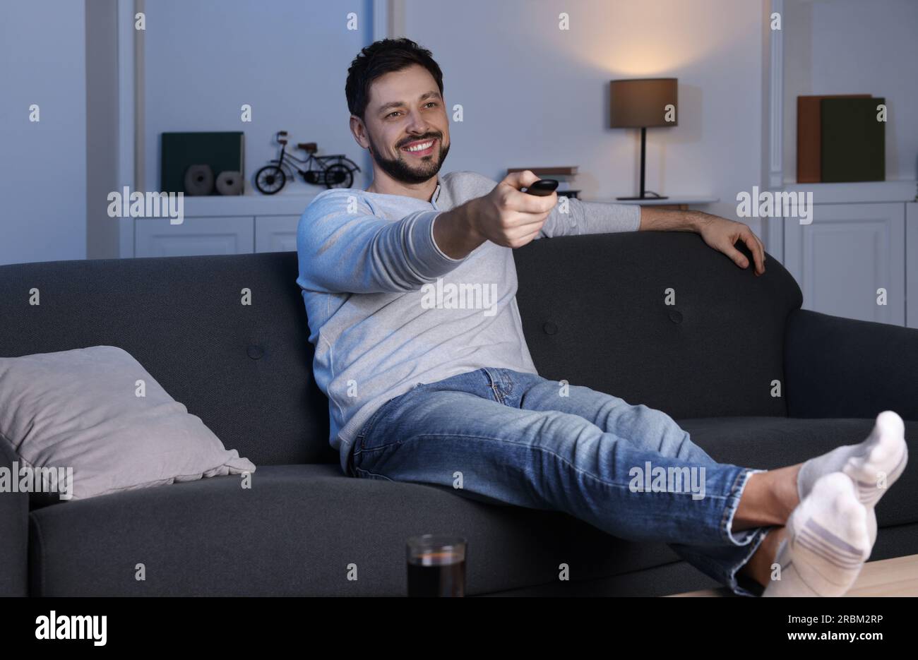 Happy man changing TV channels with remote control on sofa at home ...