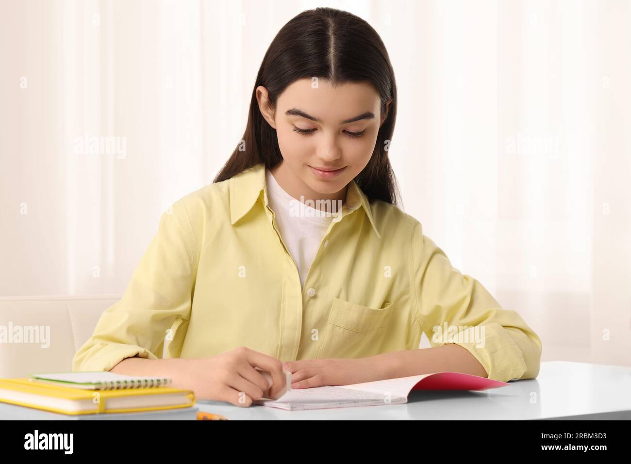 Teenage girl erasing mistake in her notebook at white desk indoors ...