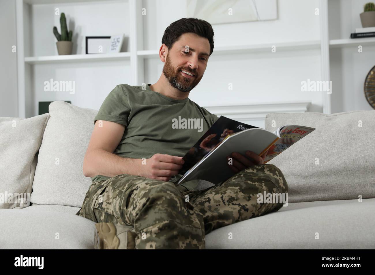 Happy soldier reading magazine on sofa in living room. Military service ...
