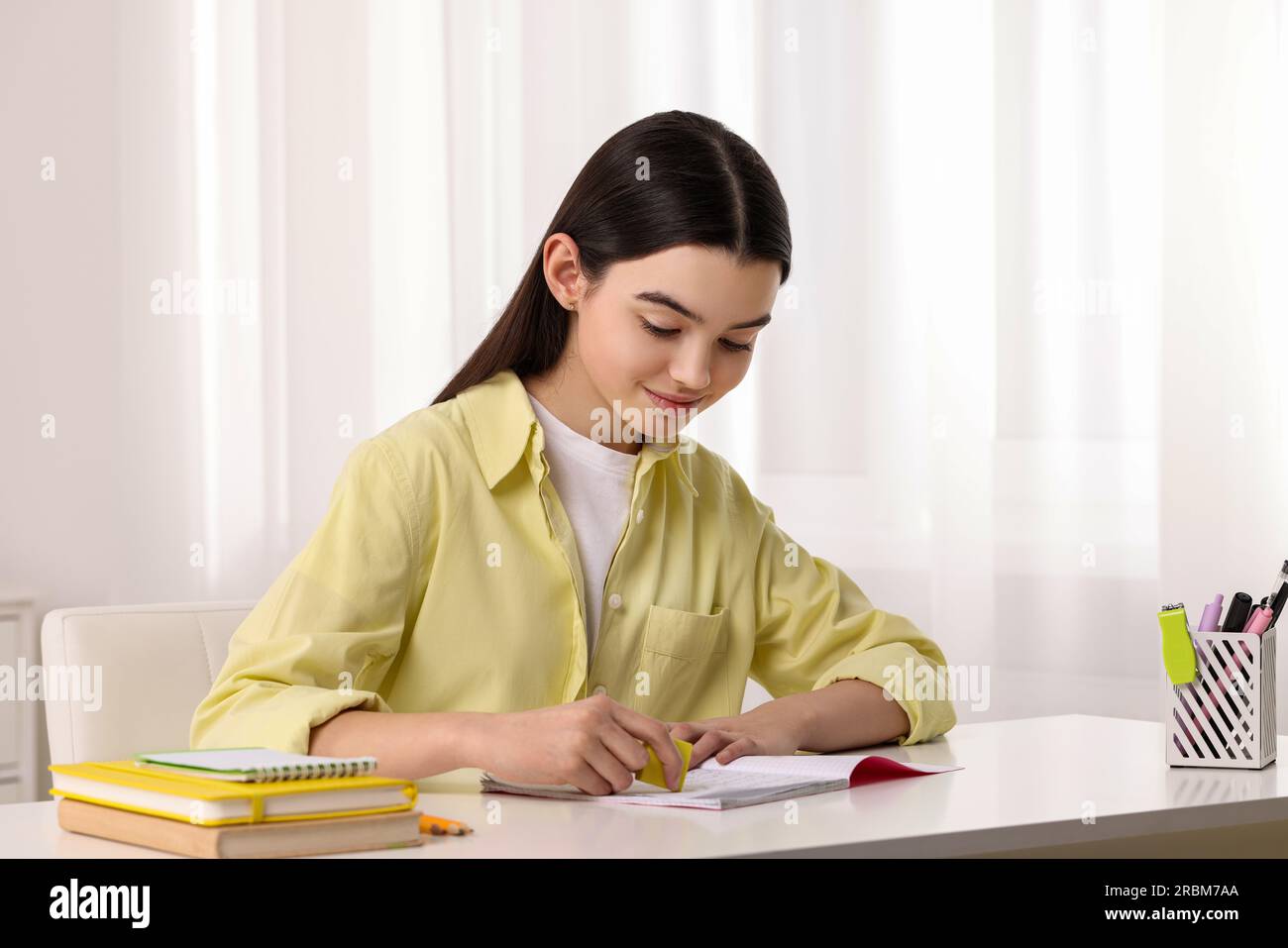 Teenage girl erasing mistake in her notebook at white desk indoors ...
