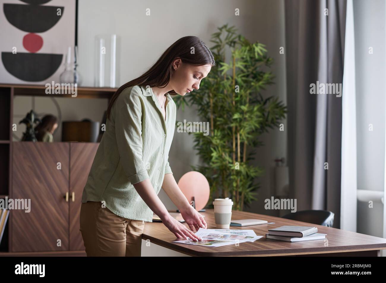 Side view of young businesswoman bending over desk and looking through ...