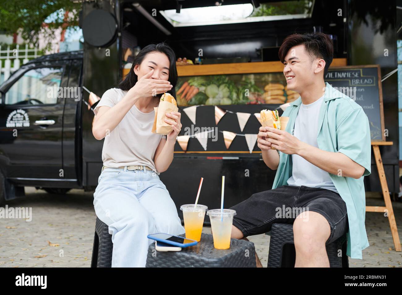 Joyful boyfriend and girlfriend eating delicious sandwiches they bought ...