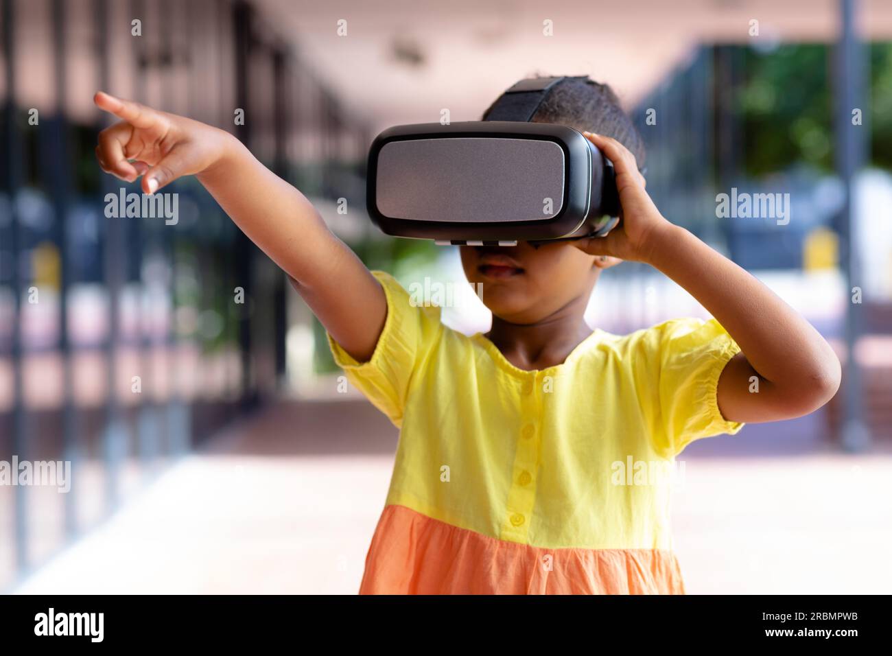 Biracial schoolgirl using vr headset pointing finger in sunny school ...