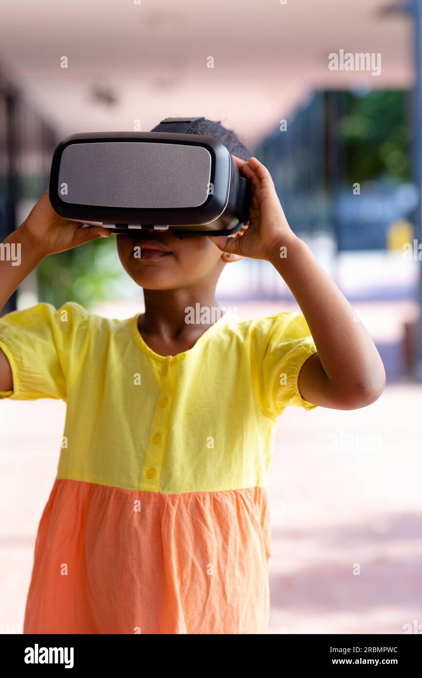 Biracial schoolgirl using vr headset in sunny school corridor, with ...