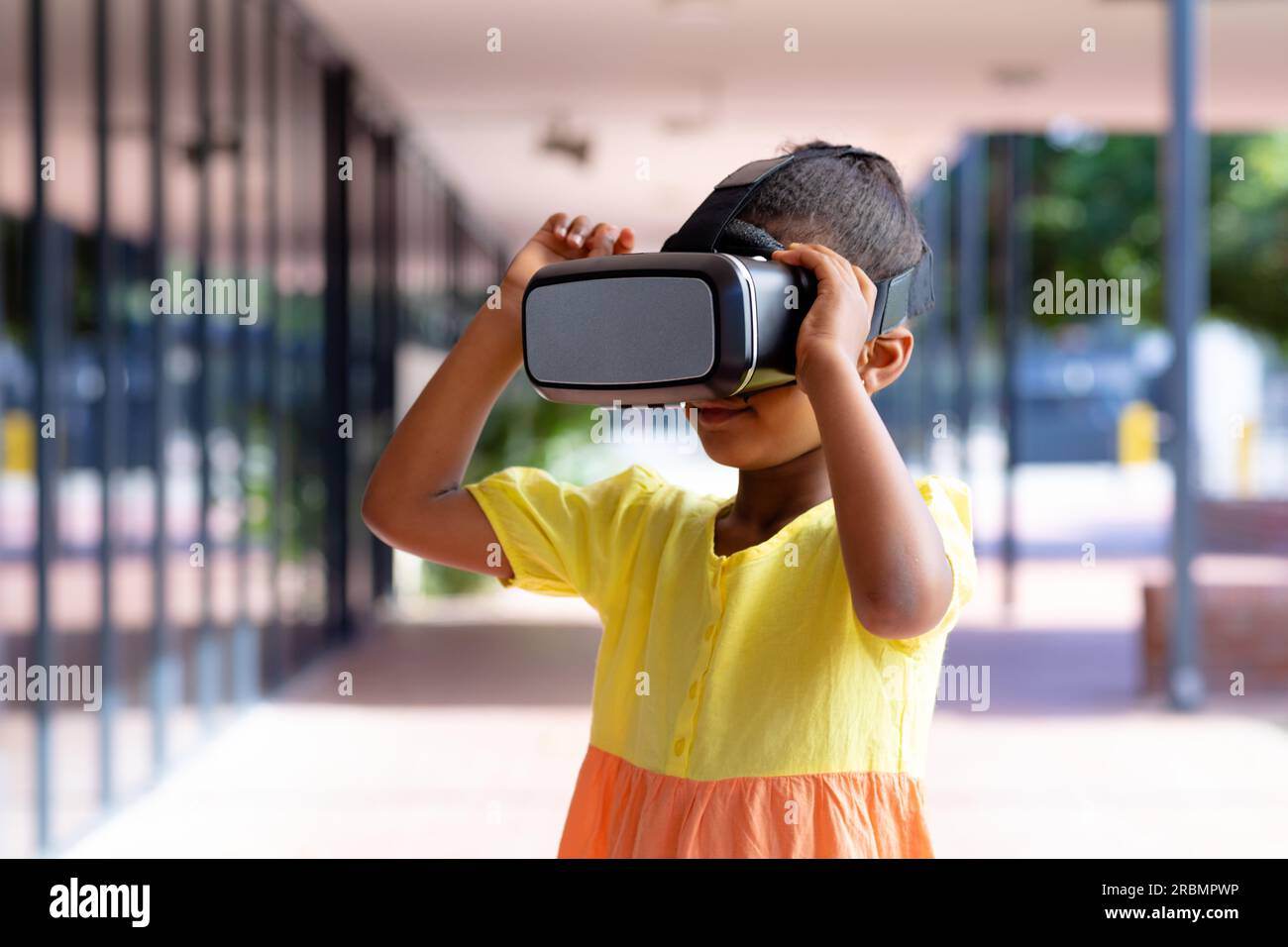 Biracial schoolgirl using vr headset in sunny school corridor with copy ...