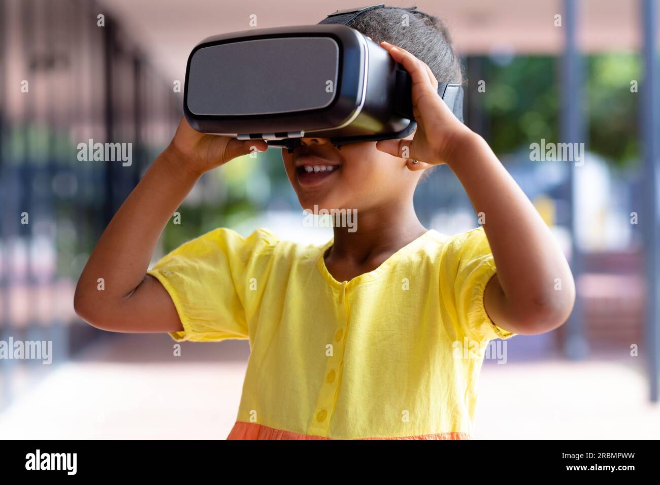 Happy biracial schoolgirl using vr headset in sunny school corridor ...