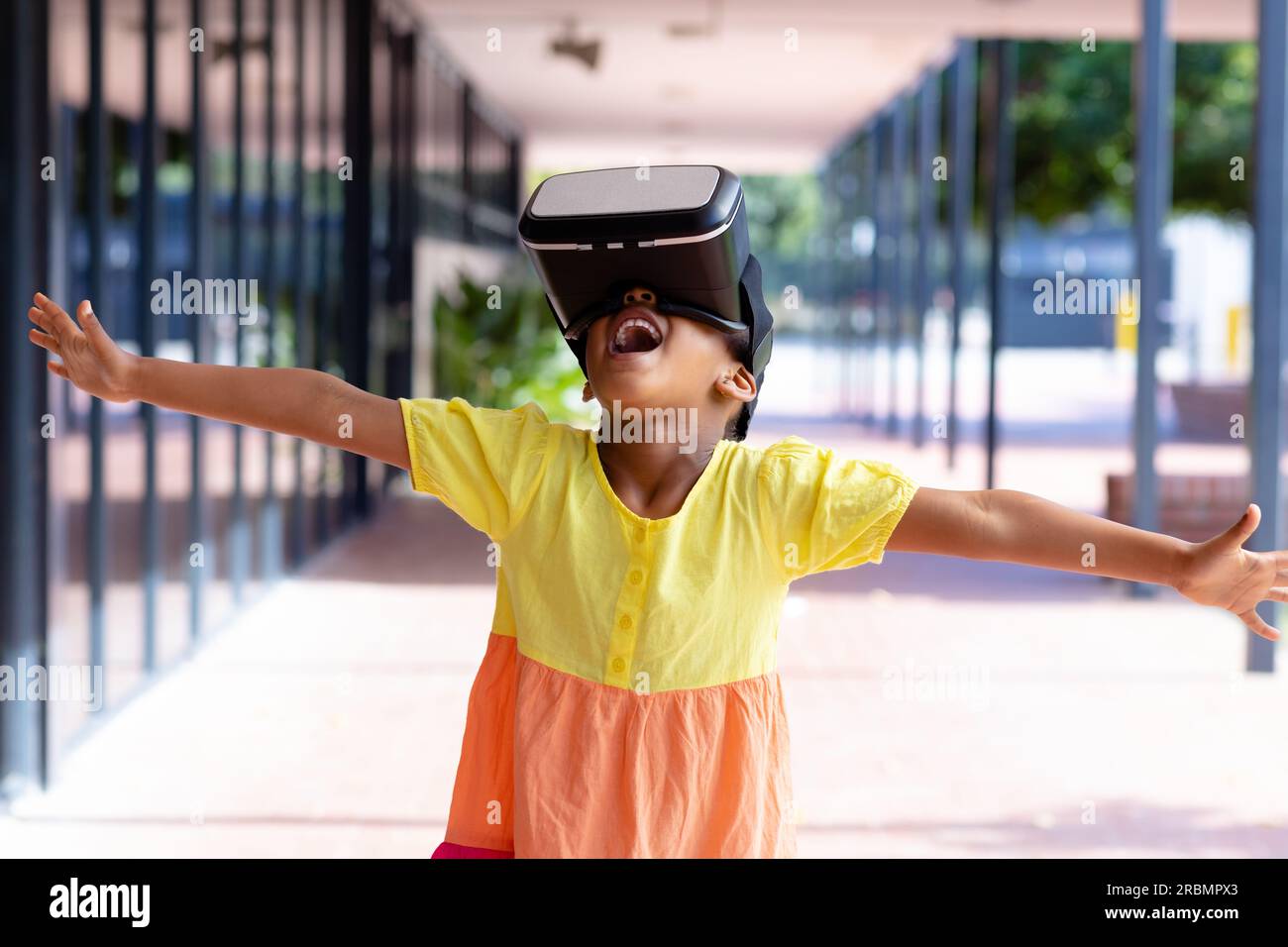Happy, biracial schoolgirl using vr headset standing in sunny school ...