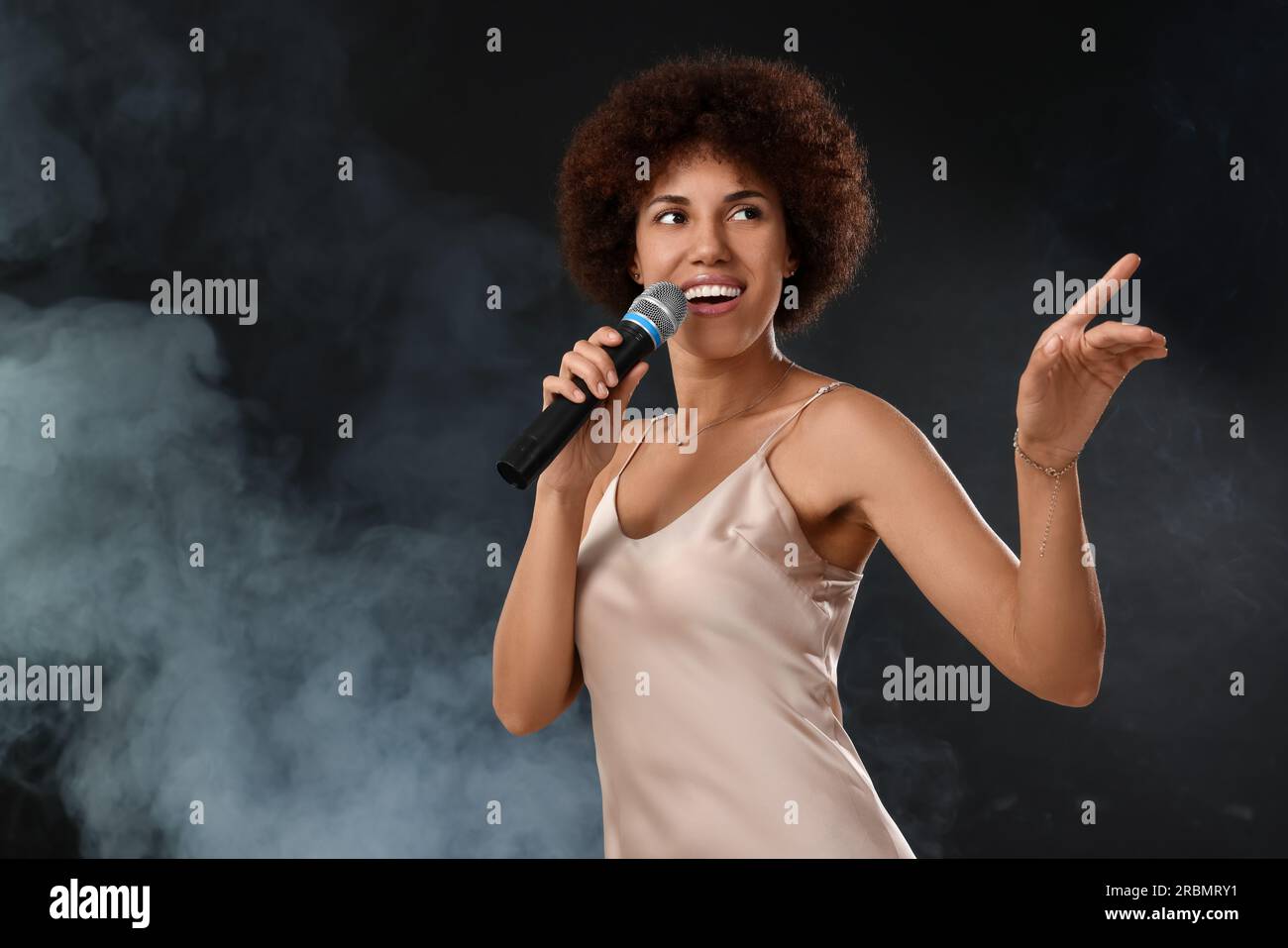 Curly young woman with microphone singing in smoke on black background ...