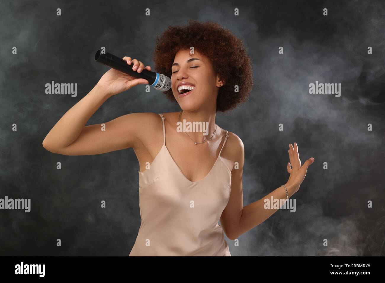 Curly young woman with microphone singing in smoke on black background ...