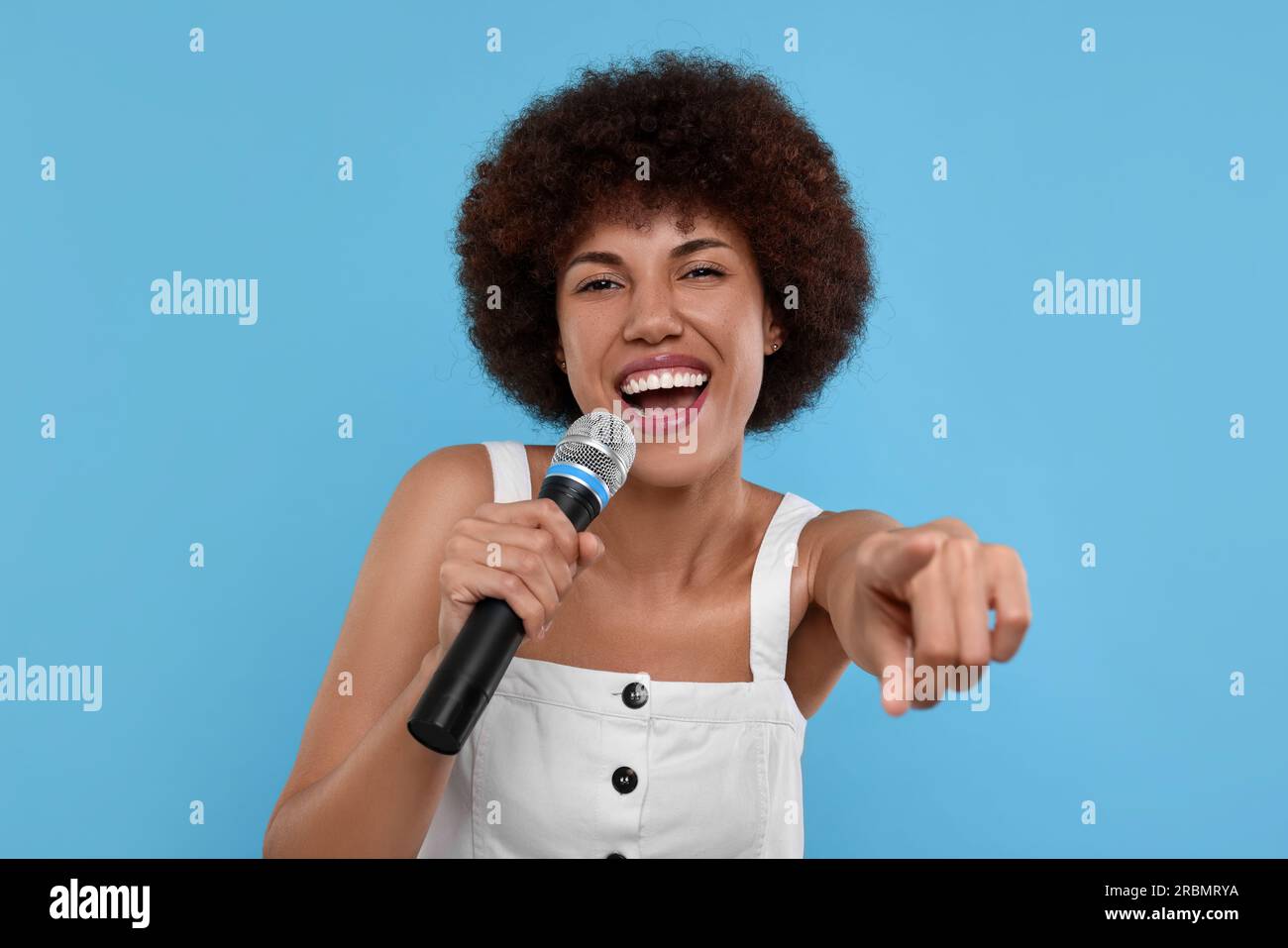 Curly young woman with microphone singing on light blue background ...