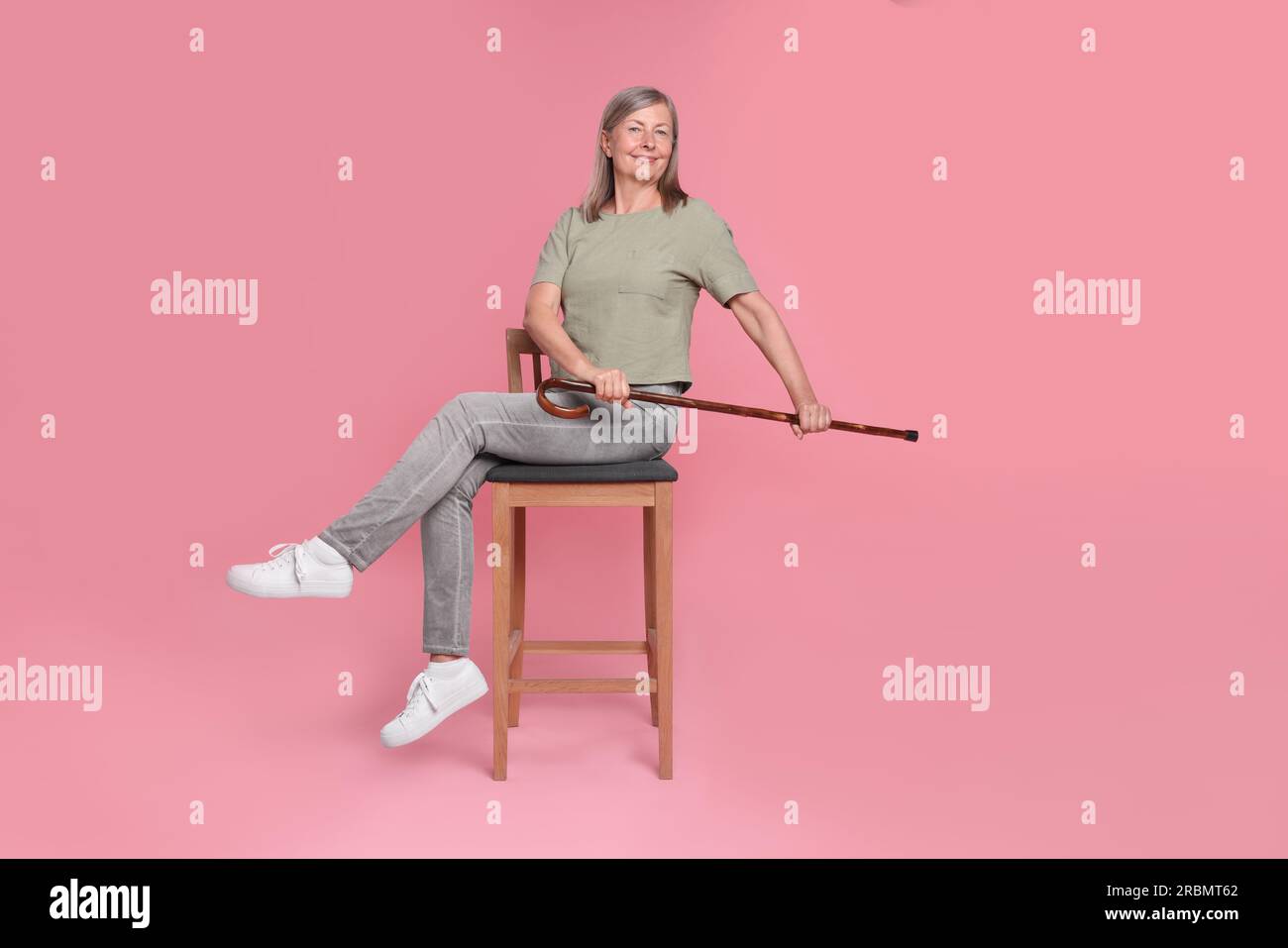 Senior woman with walking cane sitting on chair against pink background ...