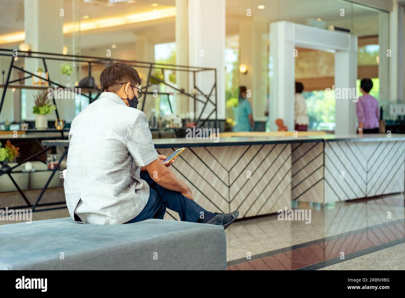 Back view of male traveler wear face mask sit to use smartphone while wait for hotel check in at ...