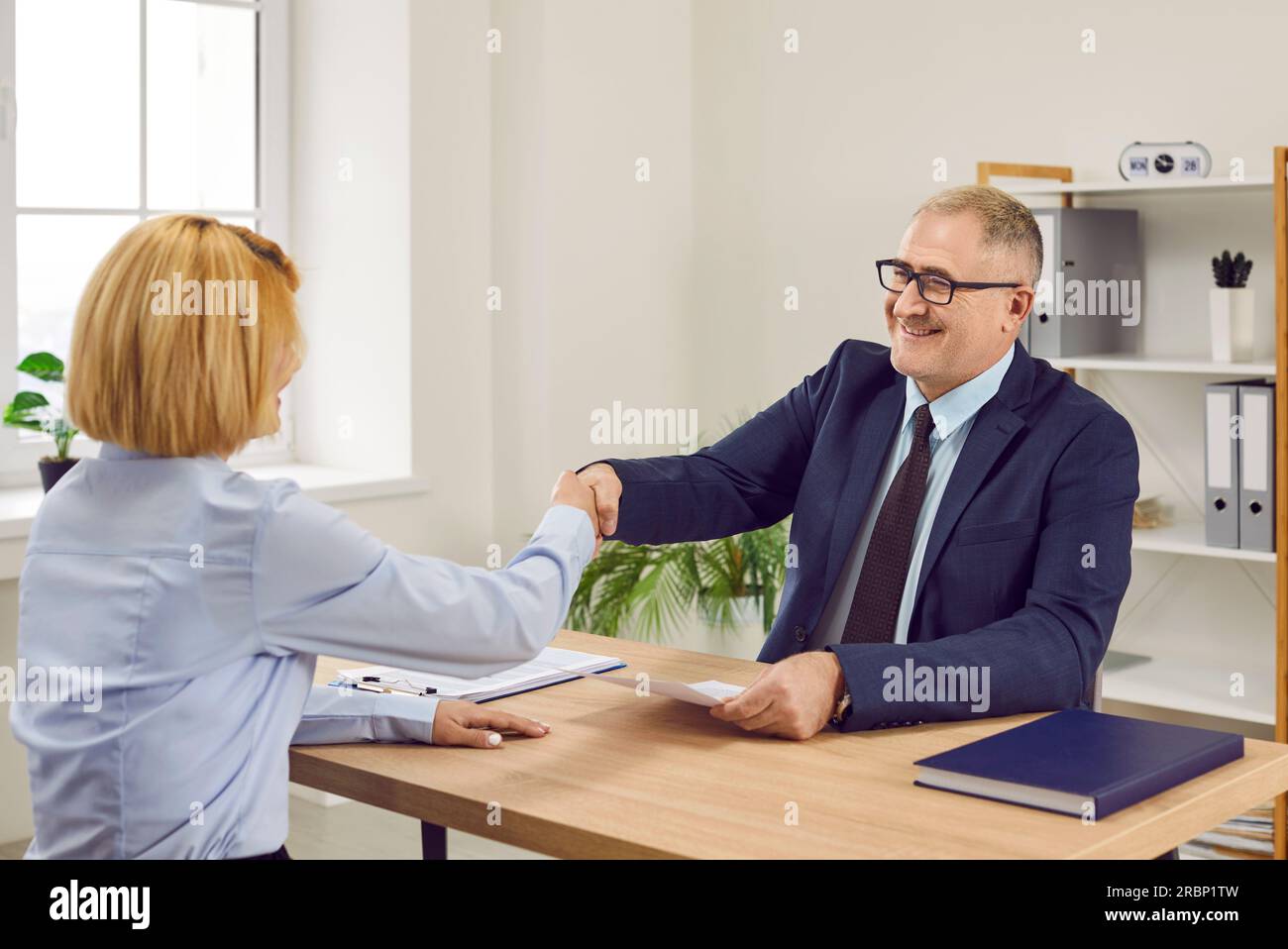 Happy senior broker sitting at office desk and exchanging handshakes ...