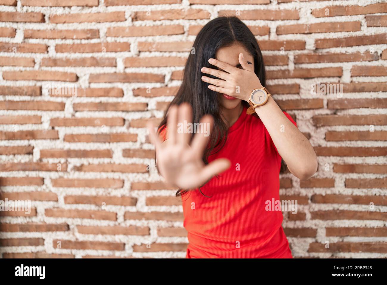Young teenager girl standing over bricks wall covering eyes with hands ...