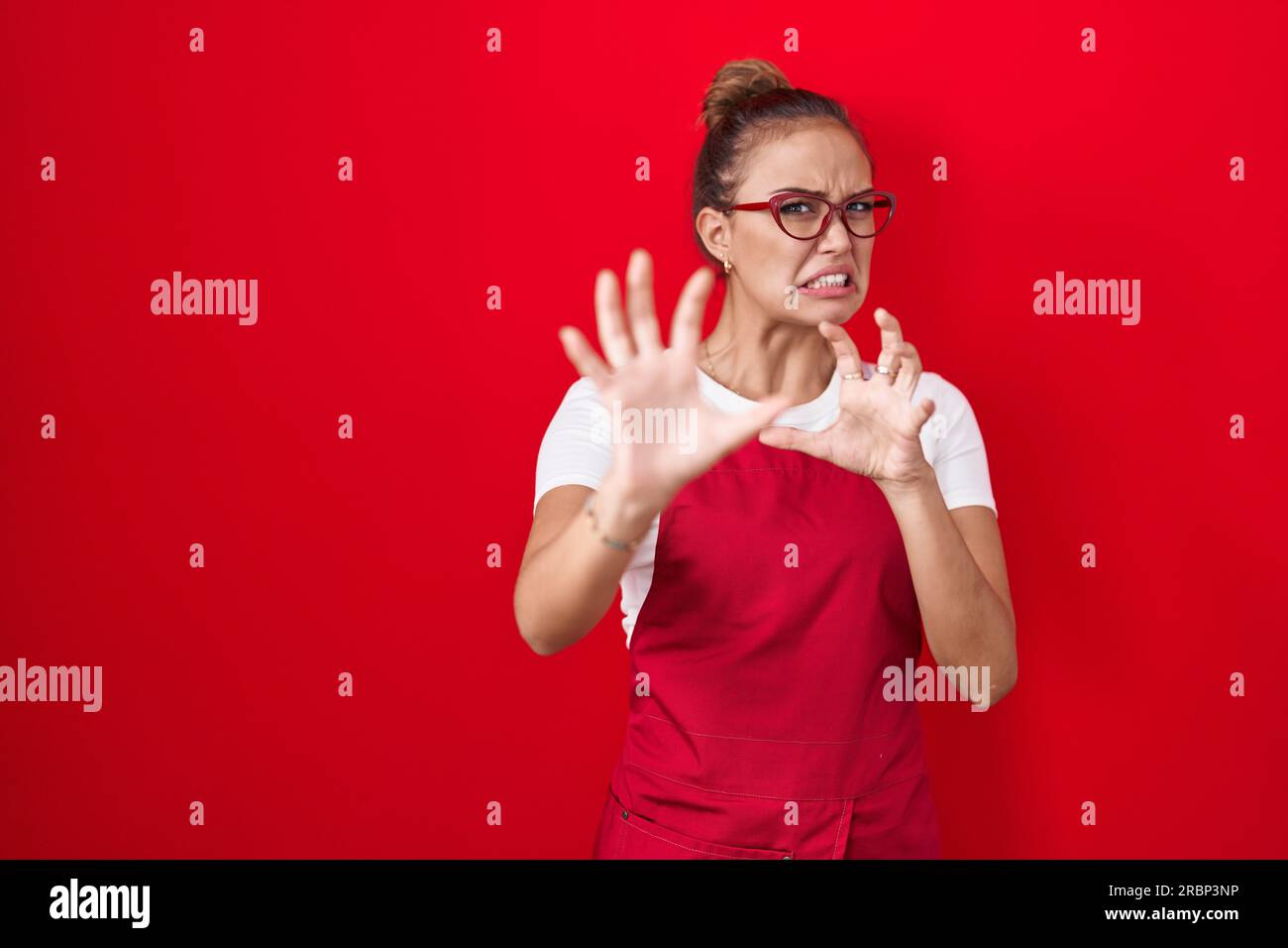 Young hispanic woman wearing waitress apron over red background afraid ...