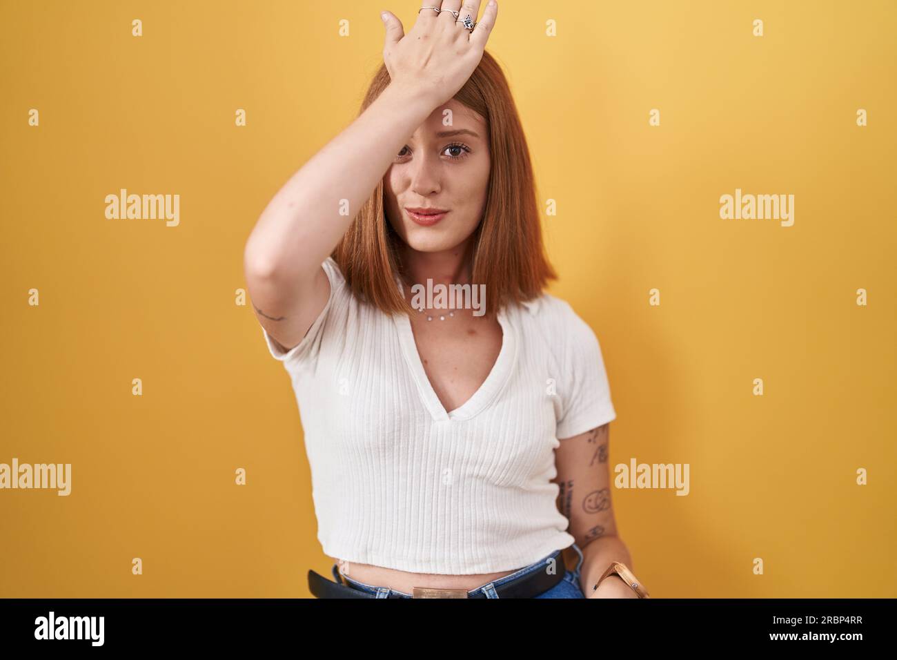 Young redhead woman standing over yellow background surprised with hand ...