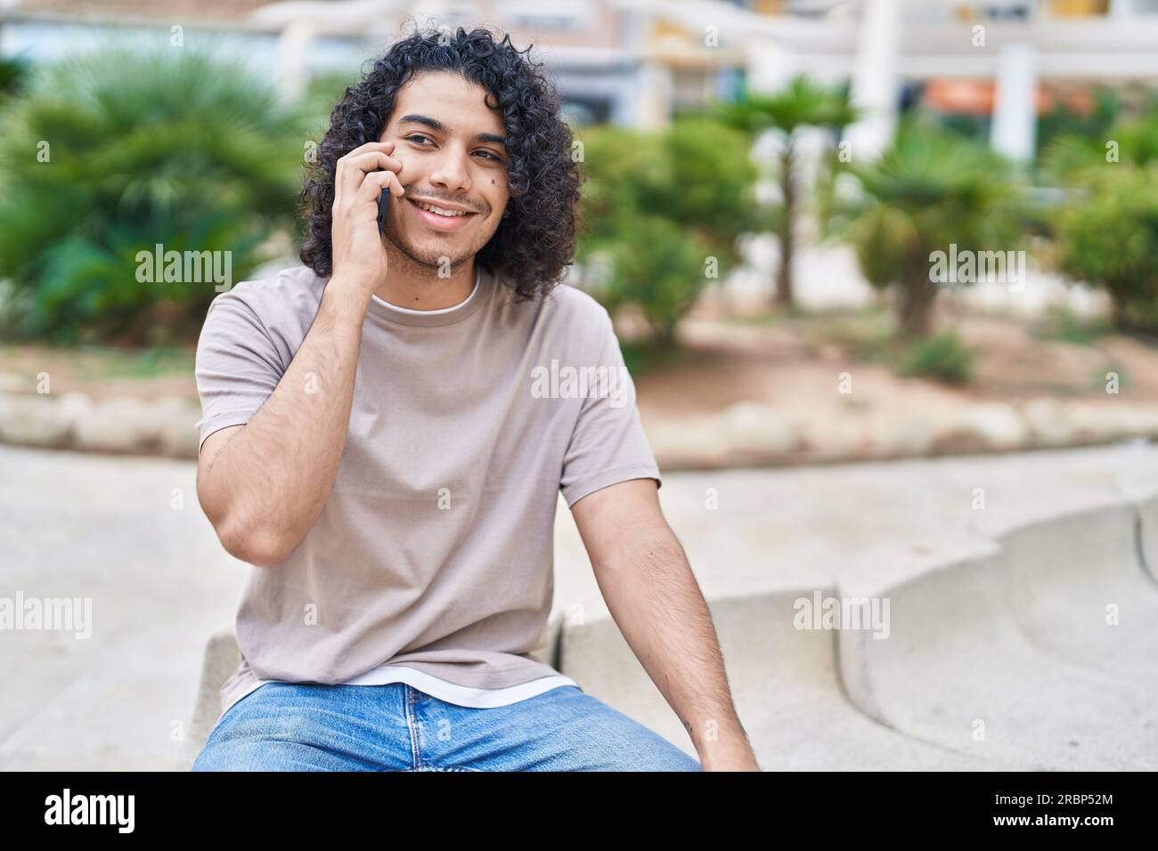 Young latin man talking on the smartphone sitting on bench at park ...