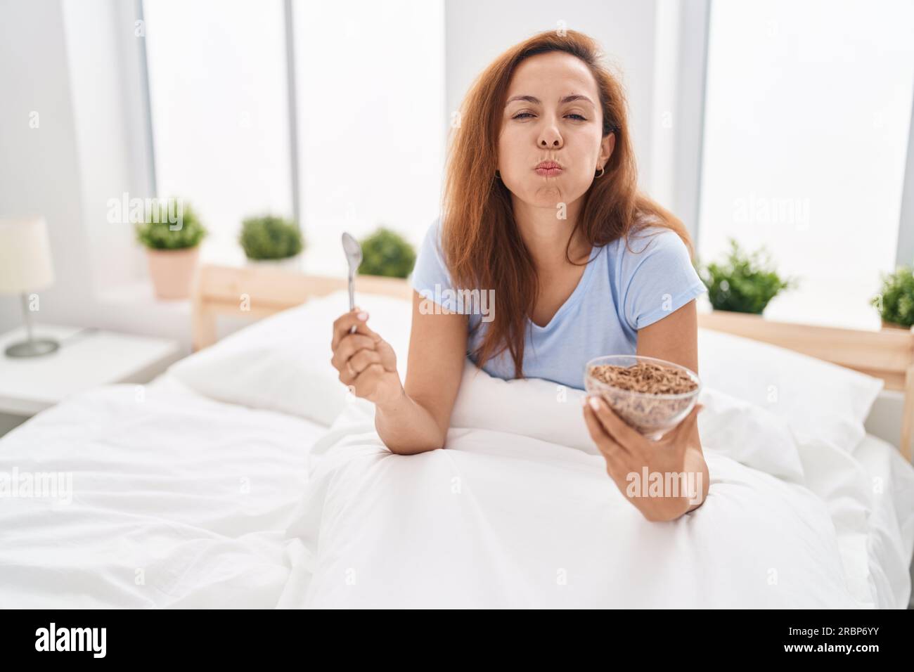 Brunette woman eating breakfast in the bed puffing cheeks with funny face. mouth inflated with ...