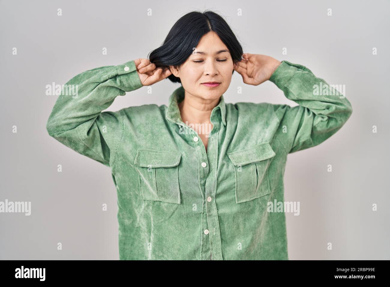 Young asian woman standing over white background smiling pulling ears ...